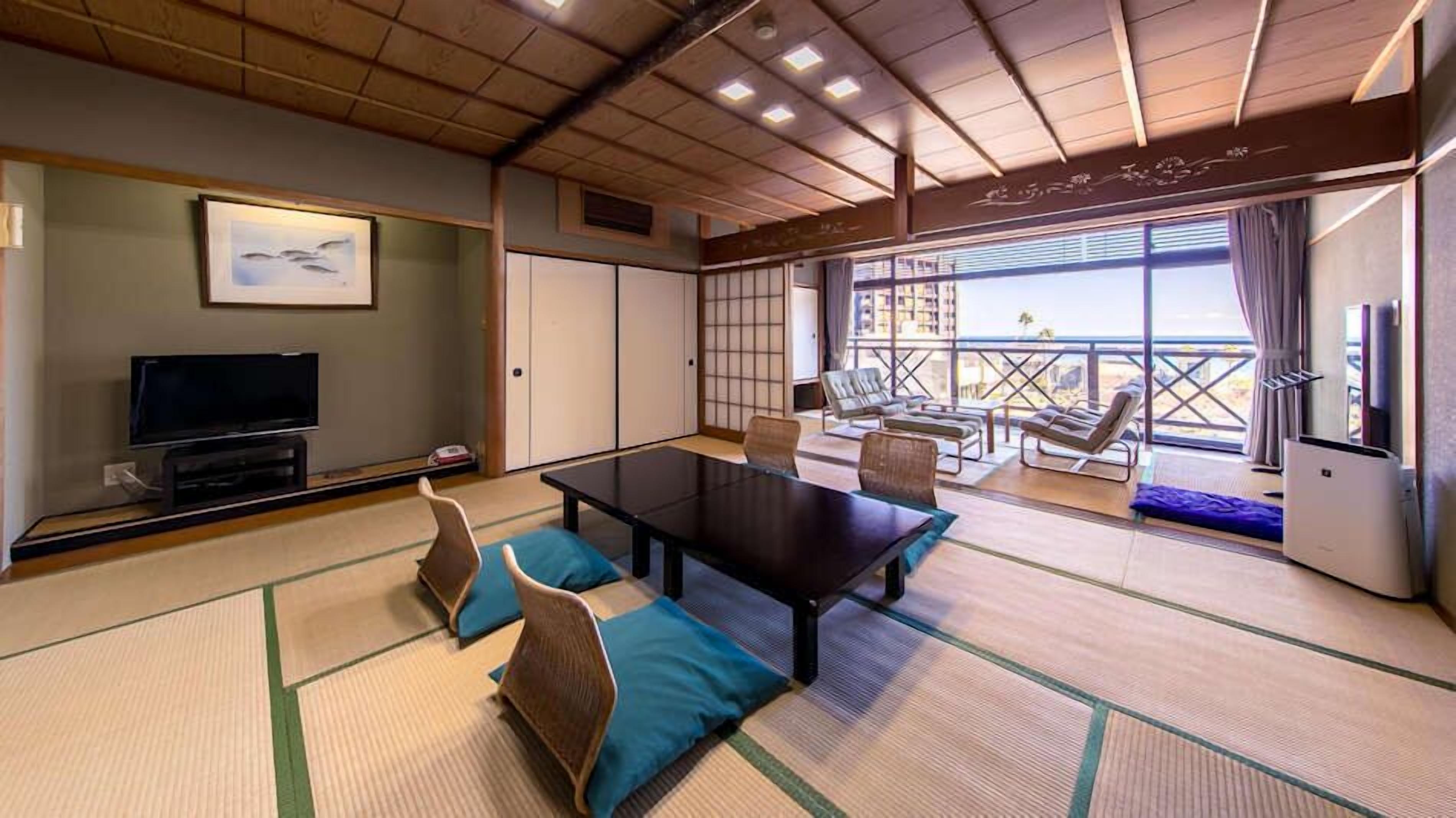 Quarto com esteiras de tatame, mesa baixa, cadeiras, televisão e varanda com espreguiçadeiras.