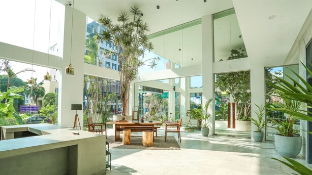 Um hall amplo com balcão de recepção, uma mesa de madeira com cadeiras e grandes vasos de plantas.
