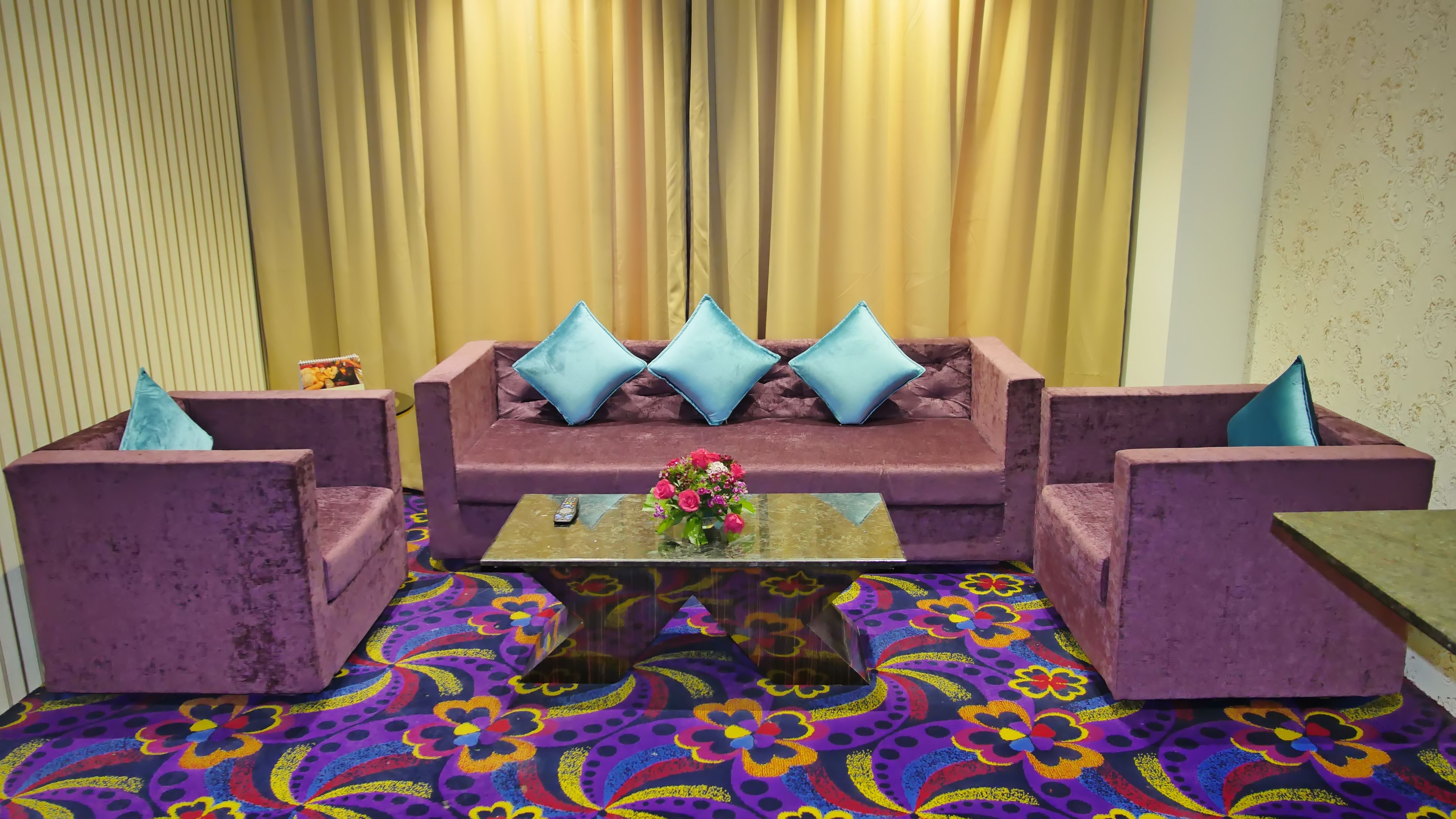 Um sofá roxo com almofadas azuis, uma mesa de centro de vidro e um controle remoto, e um tapete colorido com padrão.