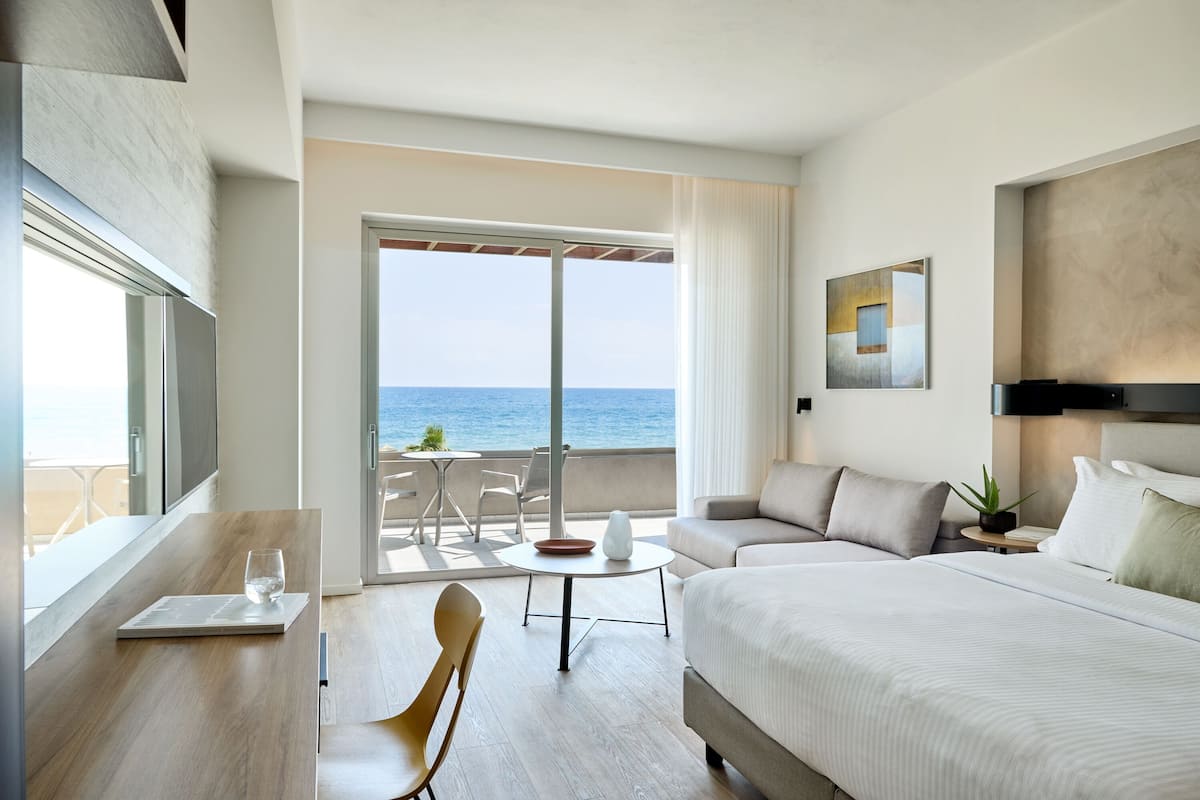 Quarto de hotel com uma cama grande, área de estar com mesa e cadeiras, e varanda com vista para o mar.