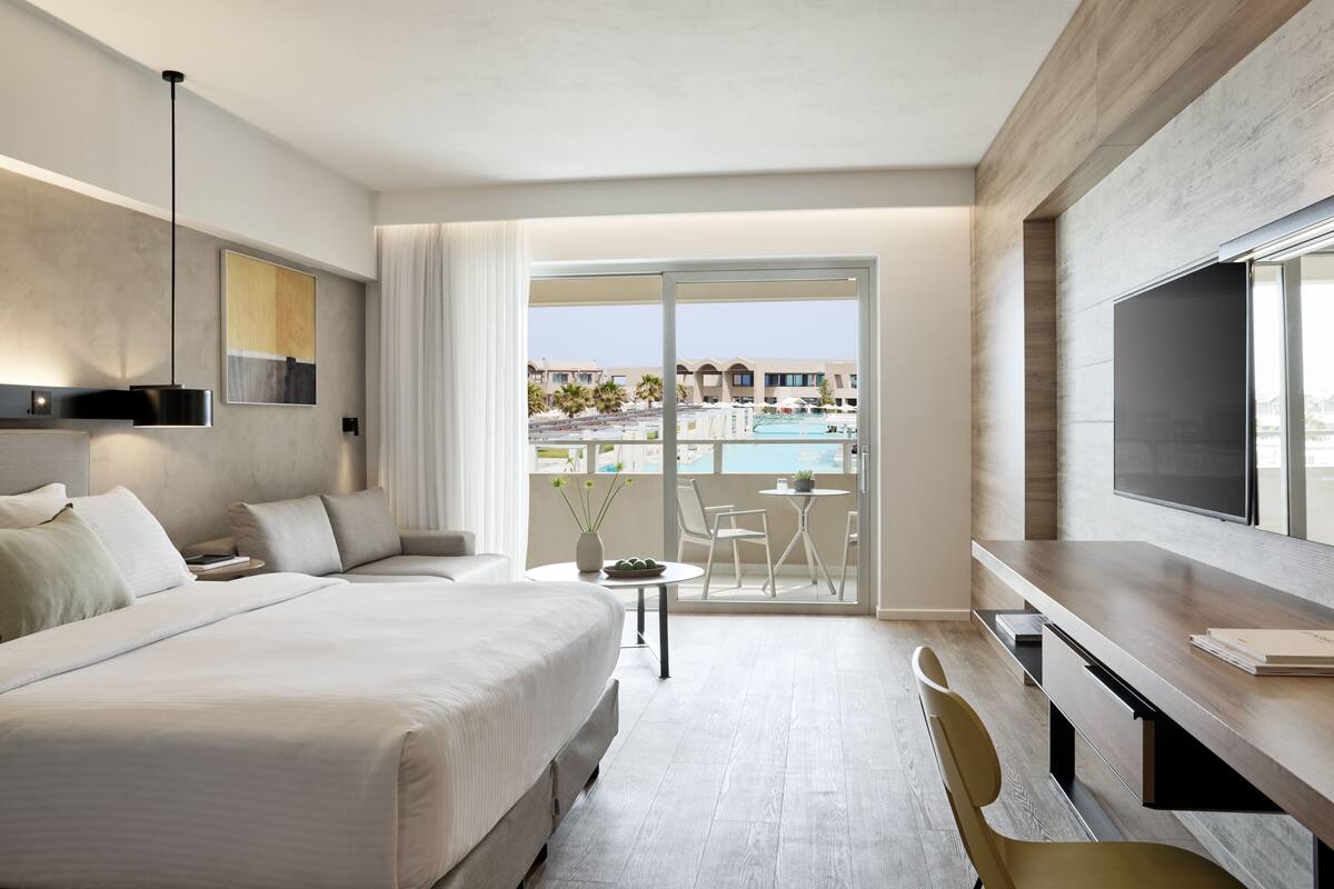 Quarto de hotel moderno com uma cama grande, uma escrivaninha, uma TV e varanda com vista para a piscina.