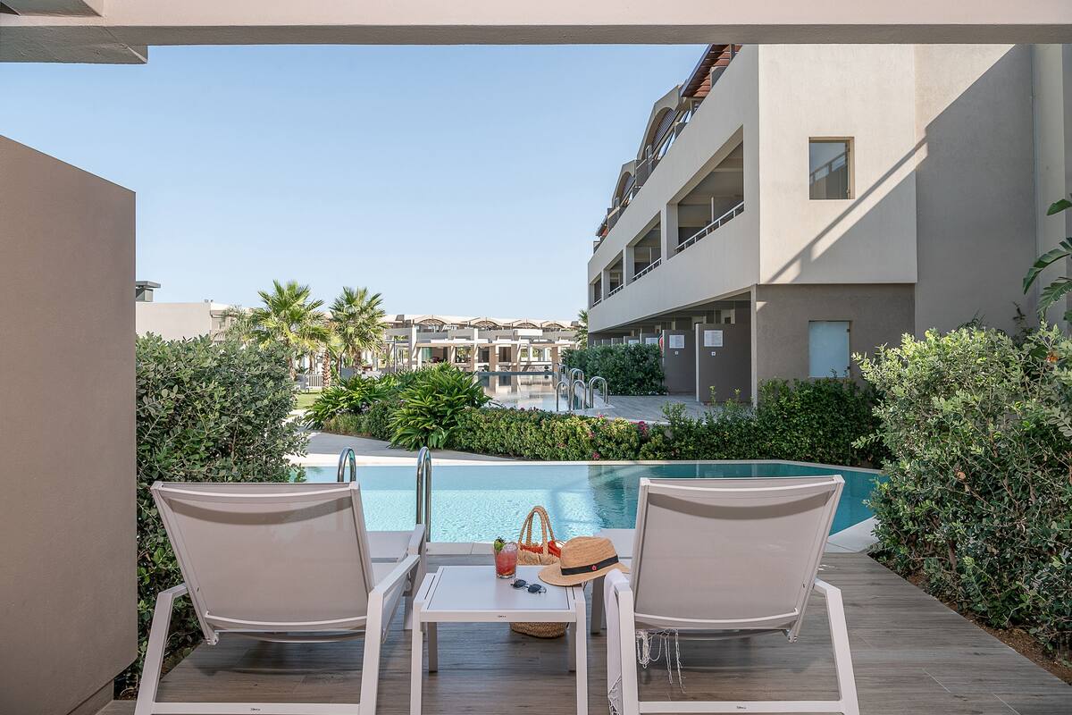 Área da piscina com duas espreguiçadeiras, uma mesa e vista para a piscina e edifícios próximos.