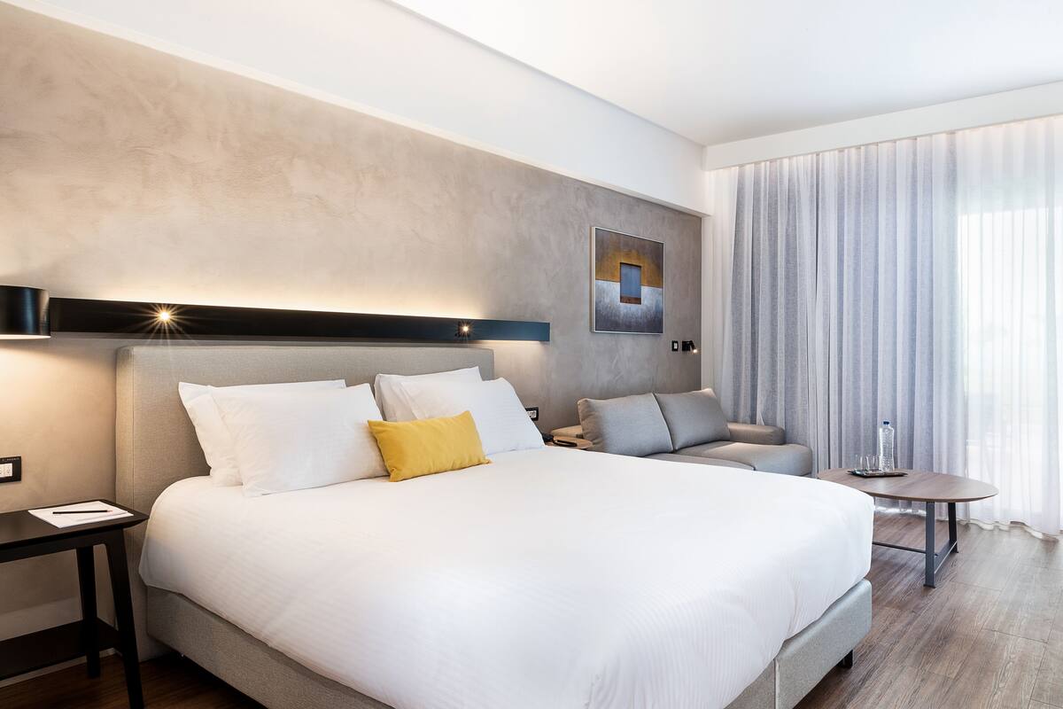 Quarto de hotel moderno com uma cama grande, uma mesa de cabeceira, um pequeno sofá, uma mesa redonda e uma janela ampla com cortinas.