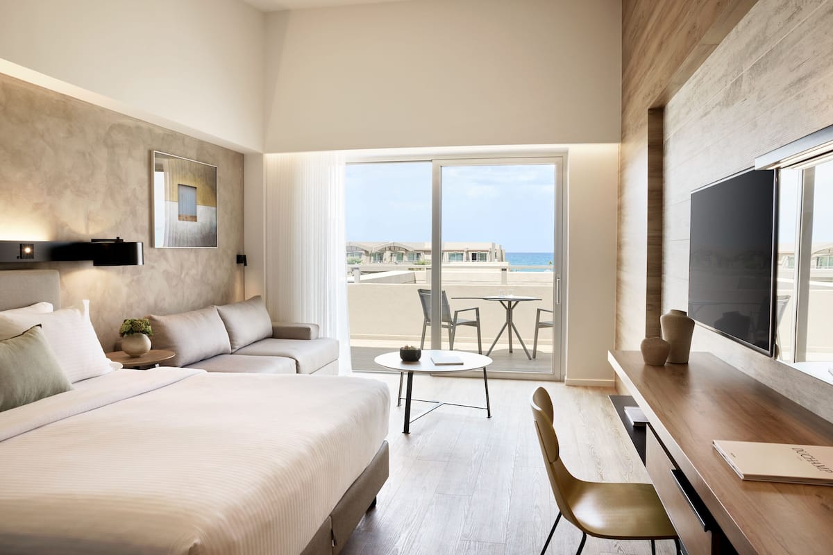 Quarto de hotel moderno com uma cama grande, uma escrivaninha, uma cadeira e vista para a praia.