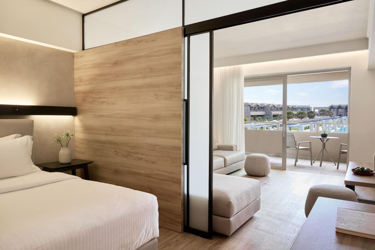 Quarto de hotel moderno com uma cama grande, parede de destaque em madeira, porta de vidro deslizante para uma varanda com assentos e vista da cidade.