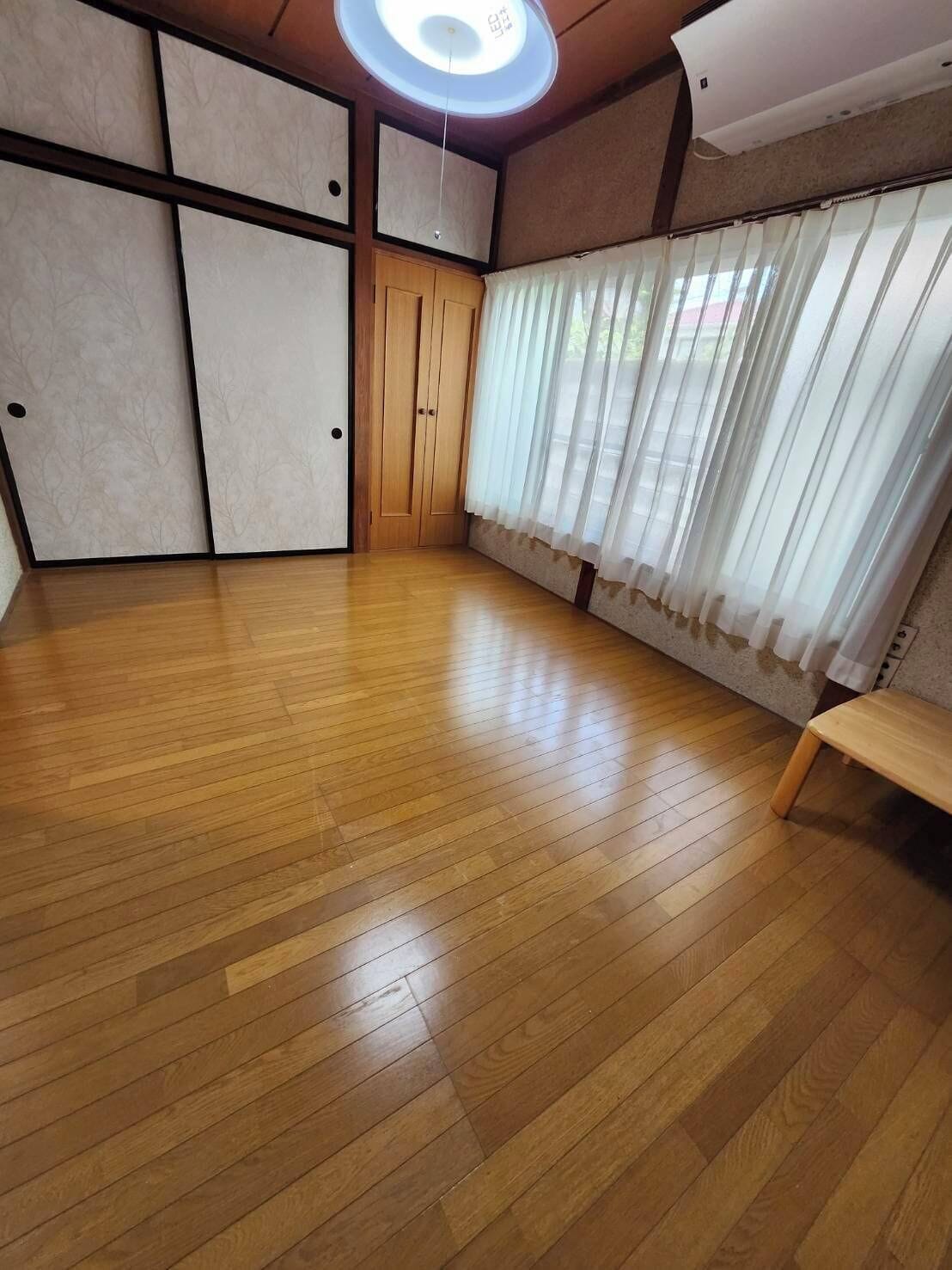 Quarto com piso de madeira, luminária no teto, janela com cortinas leves e um banco de madeira.
