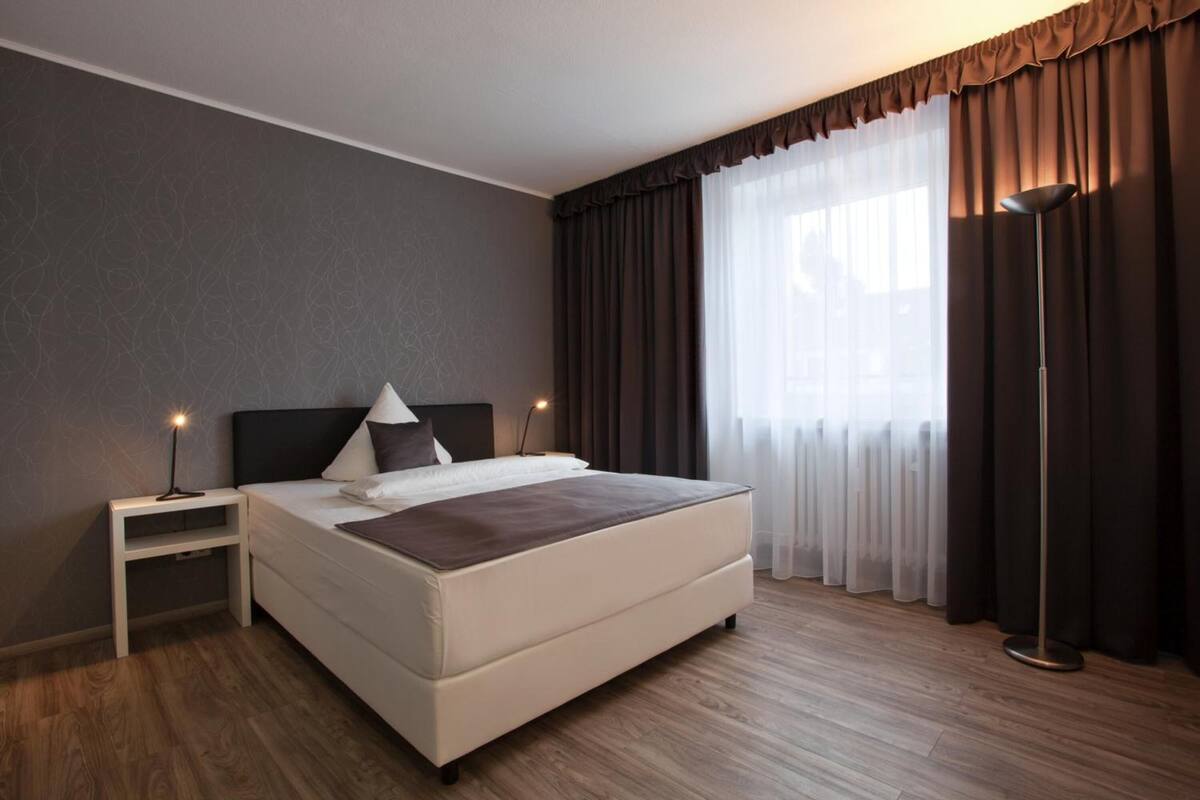 Quarto de hotel moderno com uma cama grande, uma mesa de cabeceira com abajur, um candeeiro de chão e uma janela com cortinas.