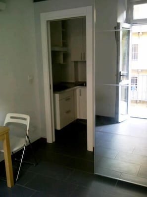 Uma cozinha com armários brancos e bancada preta, uma cadeira branca e uma porta que dá para uma área externa.