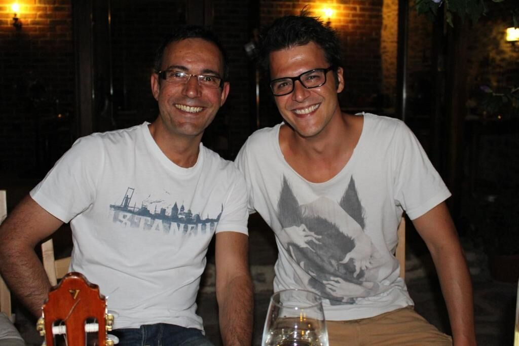Dois homens sentados à mesa, com um violão e um copo d'água.