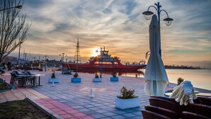Um calçadão à beira-mar com áreas de estar, uma estátua e um barco vermelho grande ao fundo.