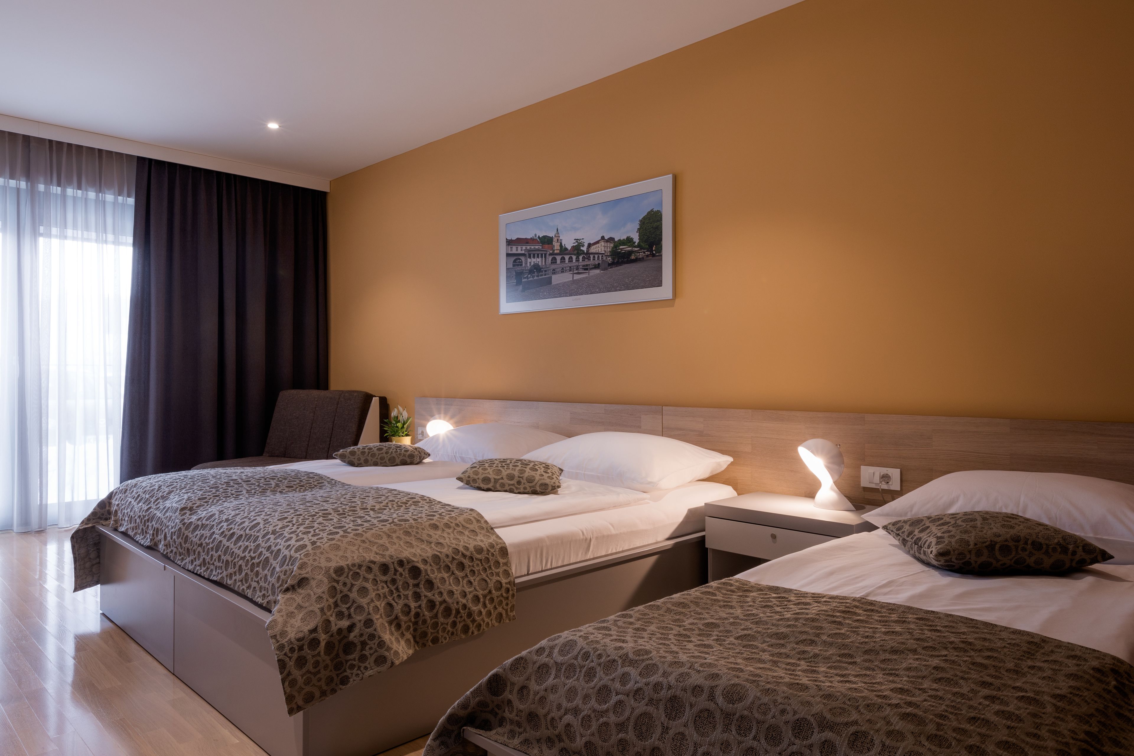 Quarto de hotel com duas camas, uma mesa de cabeceira, um abajur, uma cadeira e um quadro na parede.