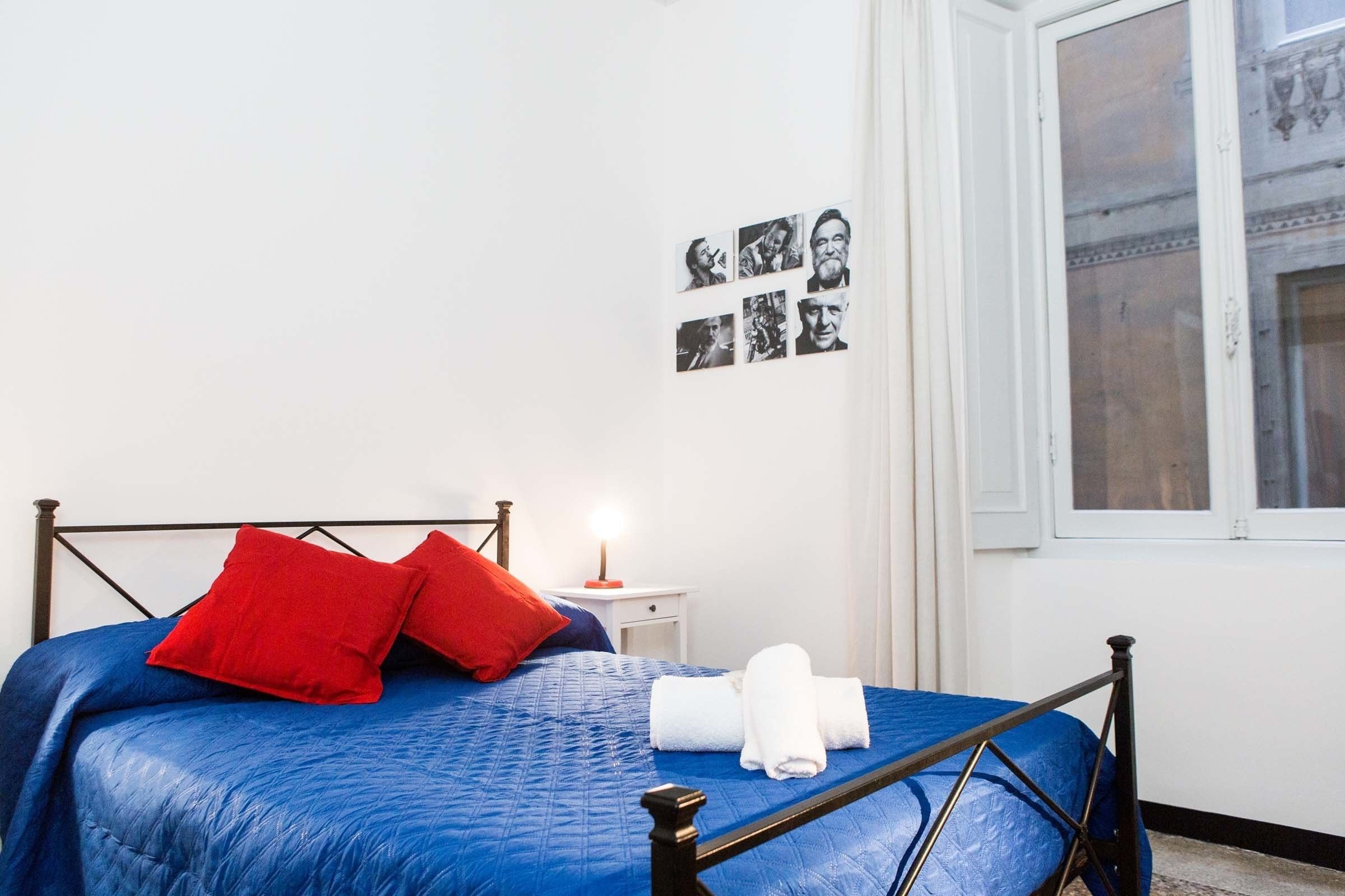 Cama bem arrumada com colcha azul e almofadas vermelhas, mesa de cabeceira com abajur e janela com vista para um prédio.