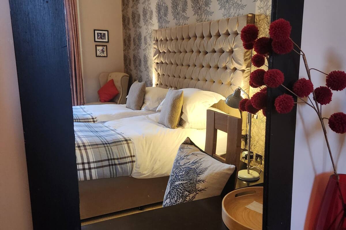 Quarto com um espelho grande refletindo o ambiente, cama com roupa branca, cabeceira estofada e um vaso de plantas vermelhas.