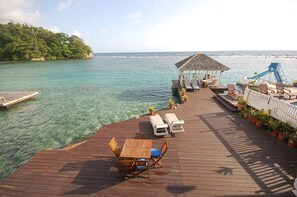 Um deck de madeira com cadeiras de descanso e uma mesa, com vista para águas turquesa cristalinas e uma ilha distante.