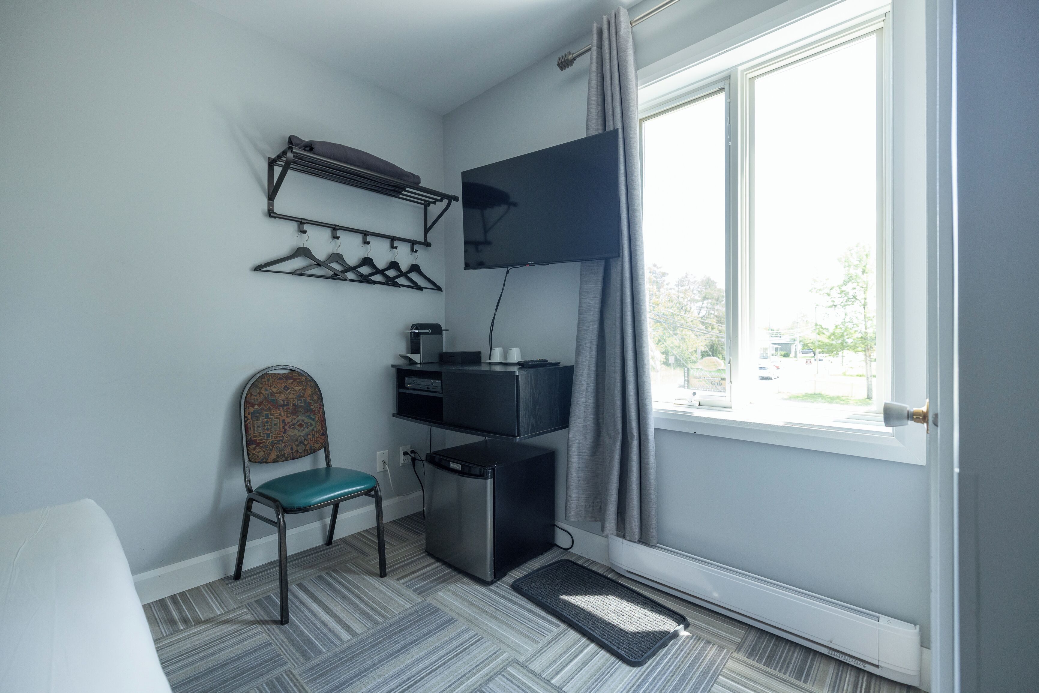 Quarto compacto com cama, poltrona, escrivaninha pequena com computador, televisão e janela com cortinas.
