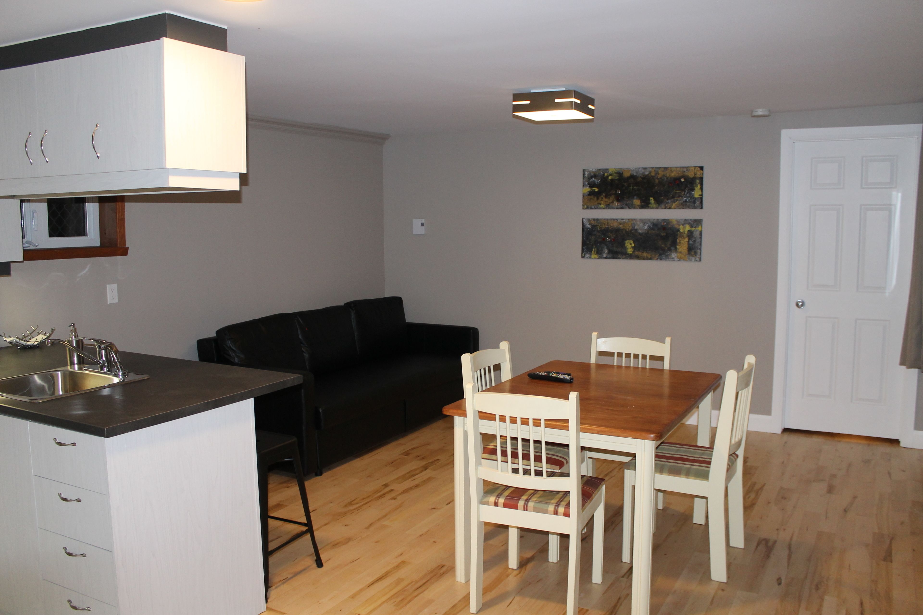 Um espaço de estar compacto com cozinha compacta, uma área de refeições com mesa e cadeiras de madeira, um sofá preto e duas pinturas abstratas na parede.