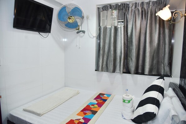 Quarto pequeno e limpo, com uma cama, televisão presa à parede, ventoinha, janela com cortinas e abajur.