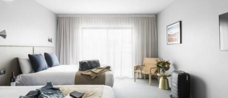 Quarto de hotel moderno com duas camas, uma escrivaninha, uma cadeira, uma mesa pequena com um vaso e uma mala.