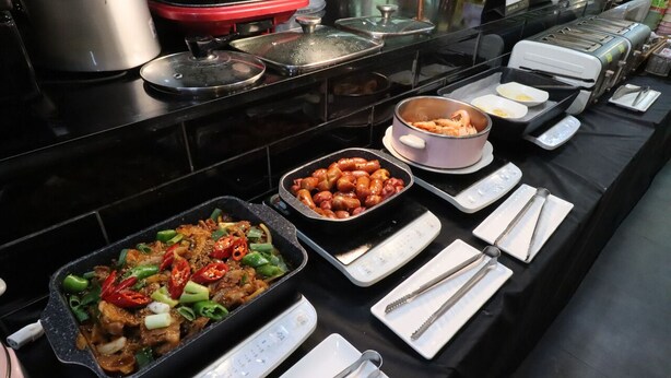 Disposição de buffet com diversos pratos, incluindo legumes refogados, salsichas e uma panela de sopa.