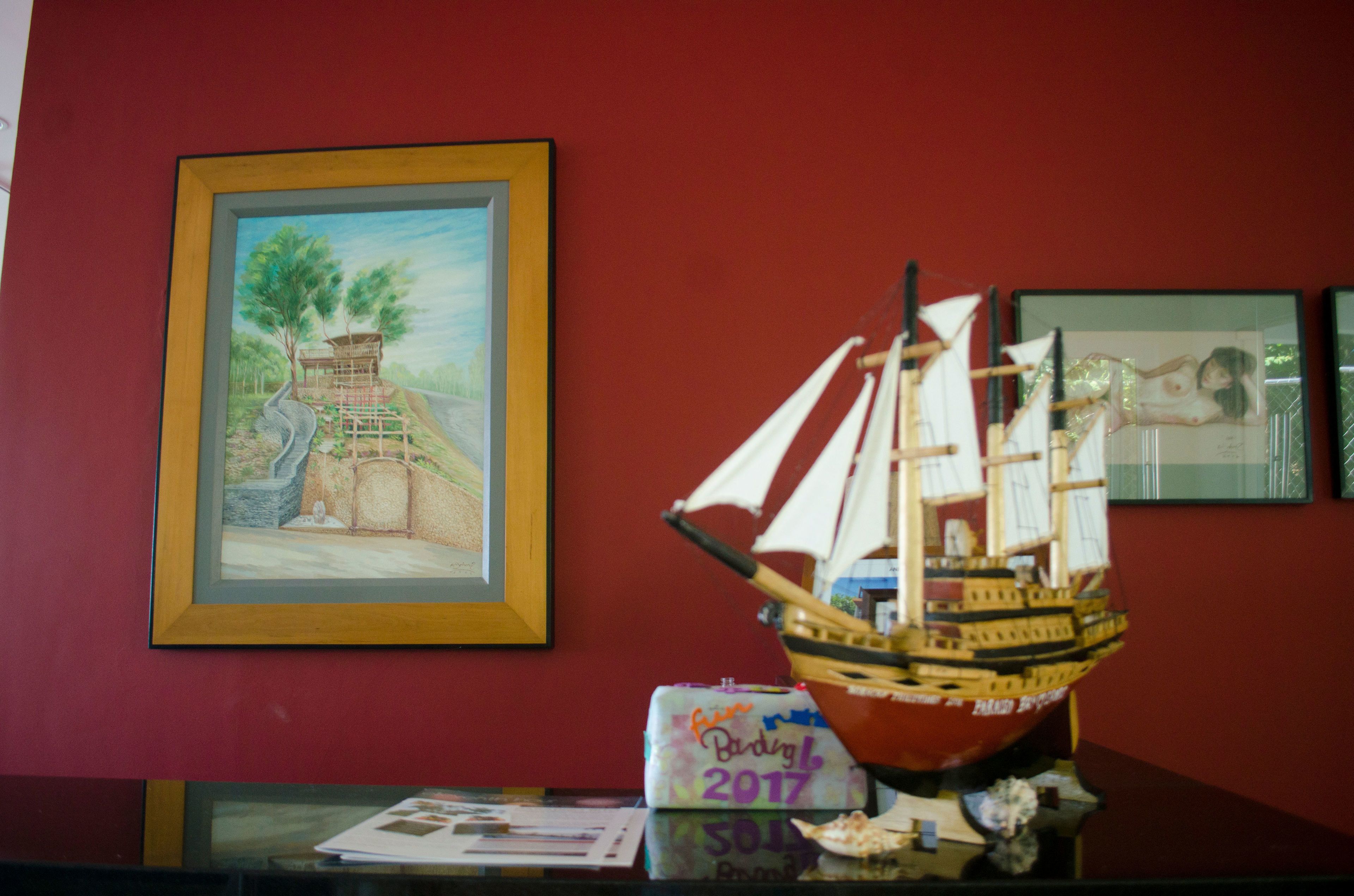 Um modelo de navio sobre uma mesa, um quadro emoldurado e uma fotografia emoldurada numa parede vermelha.