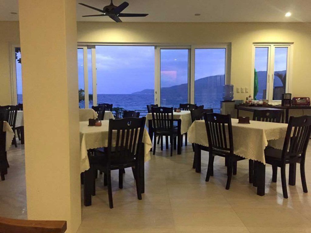 Um restaurante com várias mesas e cadeiras, janelas amplas com vista para o mar e um ventilador de teto.