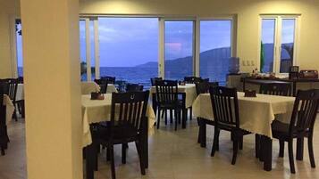 Um restaurante com várias mesas e cadeiras, janelas amplas com vista para o mar e um ventilador de teto.