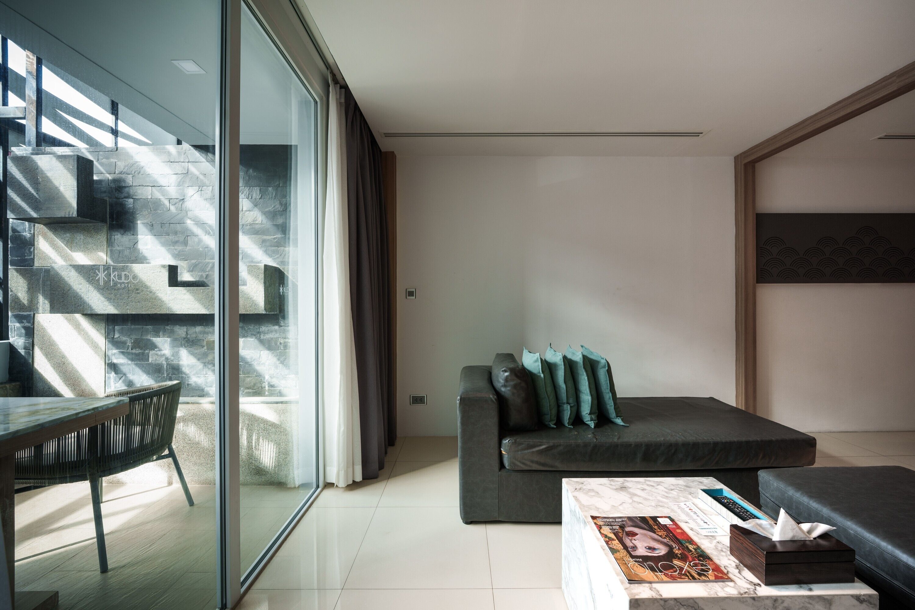 Uma sala de estar moderna com parede de vidro, sofá com almofadas, mesa de centro com revista e um banco de madeira.