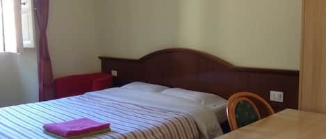 Uma cama de solteiro com colcha listrada, um travesseiro vermelho e uma toalha rosa dobrada sobre a cama. Há uma mesinha de cabeceira de madeira com uma luminária, uma cadeira e uma escrivaninha de madeira com cadeira. O quarto tem uma janela com cortinas, um candeeiro fixado na parede e um quadro acima da cama.