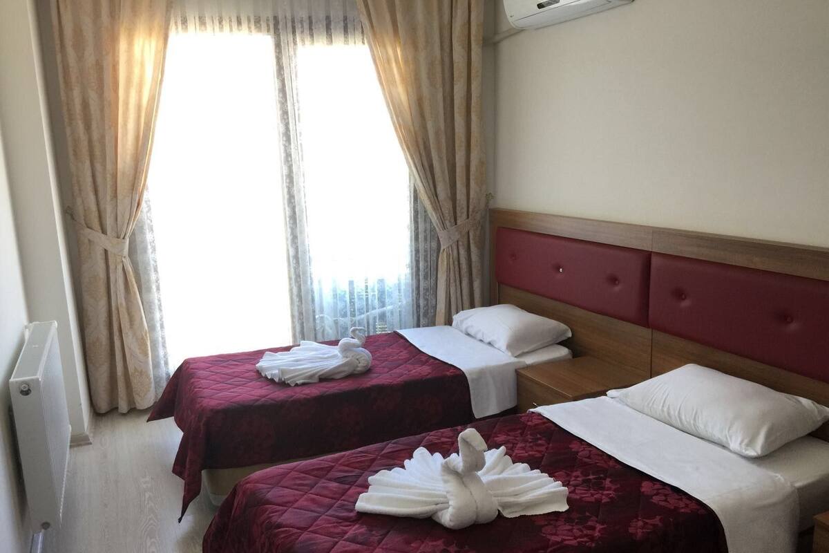 Quarto de hotel com duas camas, cada uma com colcha vermelha e almofadas brancas. Há duas toalhas brancas em formato de cisne sobre as camas. O quarto possui cabeceira de madeira e uma janela com cortinas.