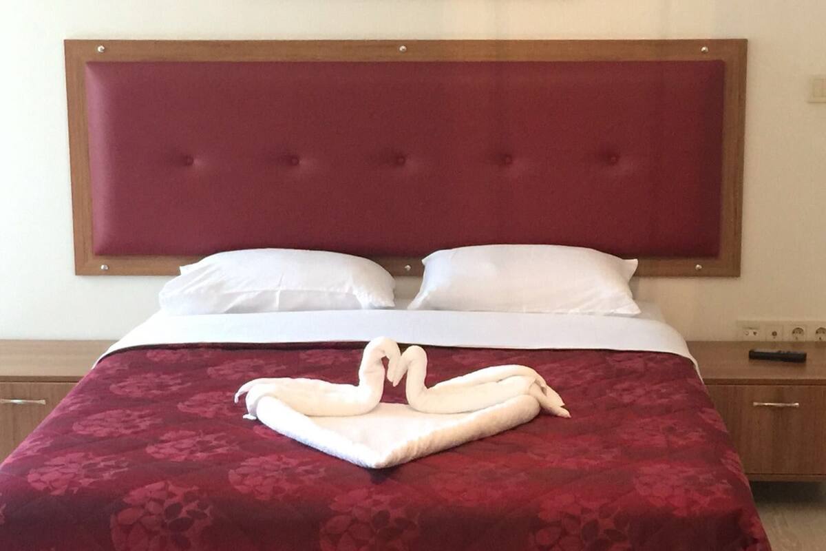 Quarto de hotel com uma cama grande, colcha vermelha com padrão floral, almofadas brancas e cabeceira de madeira.