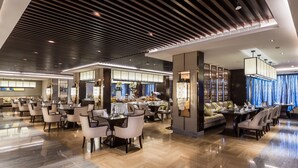 Um hall de hotel espaçoso com design moderno, apresentando uma grande área de estar com mesas e cadeiras, uma parede decorativa com padrões intrincados e um teto com um design de ripas único.