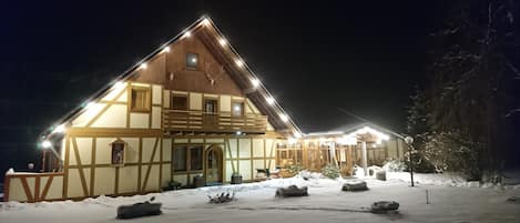 Um edifício grande e iluminado, com telhado inclinado, várias janelas e chão coberto de neve.