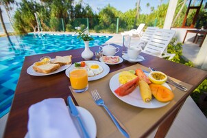 Uma mesa de café da manhã com torradas, ovos e frutas, um vaso de flores e vista para uma piscina e vegetação.