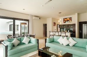 Uma sala de estar moderna com sofás azul-turquesa e uma mesa de centro de vidro, uma área de jantar com uma cozinha ao fundo, e uma grande janela com vista para o exterior.