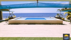 Piscina com vista para o oceano, cercada por plantas em vasos e uma área arborizada.