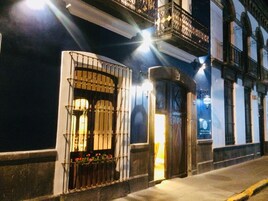 Fachada de um edifício à noite, com entrada em destaque, varanda com grades ornamentadas e um poste de luz iluminando a área.