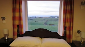 Uma cama com almofadas brancas e um edredom estampado em vermelho e preto, com duas mesas de cabeceira com abajures, em um quarto com cortinas xadrez e vista para uma paisagem verde pela janela.