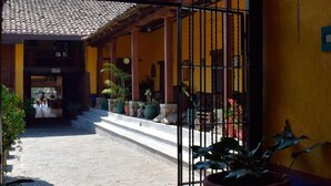 Um pátio com piso de azulejos, plantas em vasos, um portão e um sino na parede.