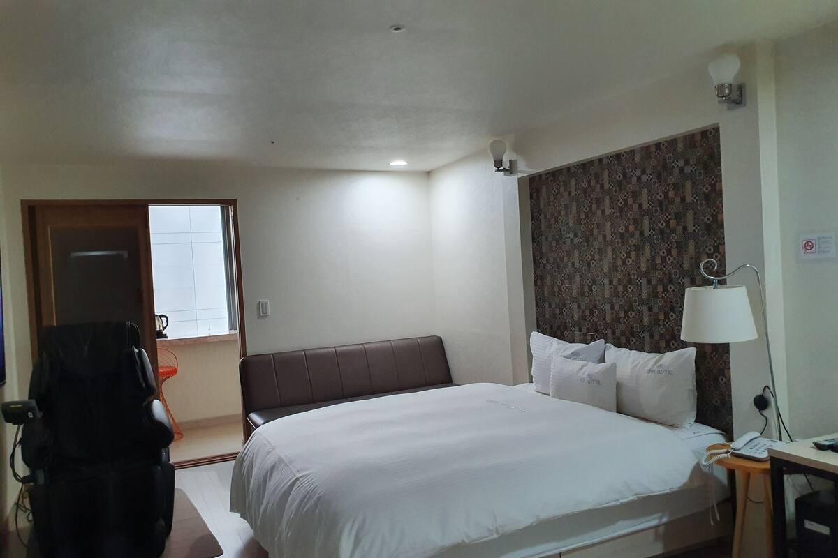Quarto de hotel com cama, escrivaninha com luminária, cadeira e poltrona de massagem.