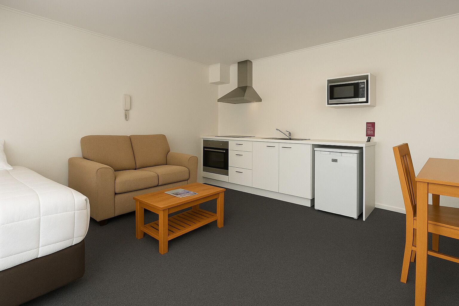 Quarto de hotel compacto com cama, sofá, pequena mesa de madeira, cozinha compacta com micro-ondas e uma cadeira.