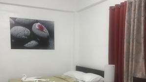 Quarto de hotel com duas camas, cada uma com colcha verde e almofadas brancas. Há uma pintura na parede acima das camas, cortina vermelha na janela e abajur branco na mesa de cabeceira.