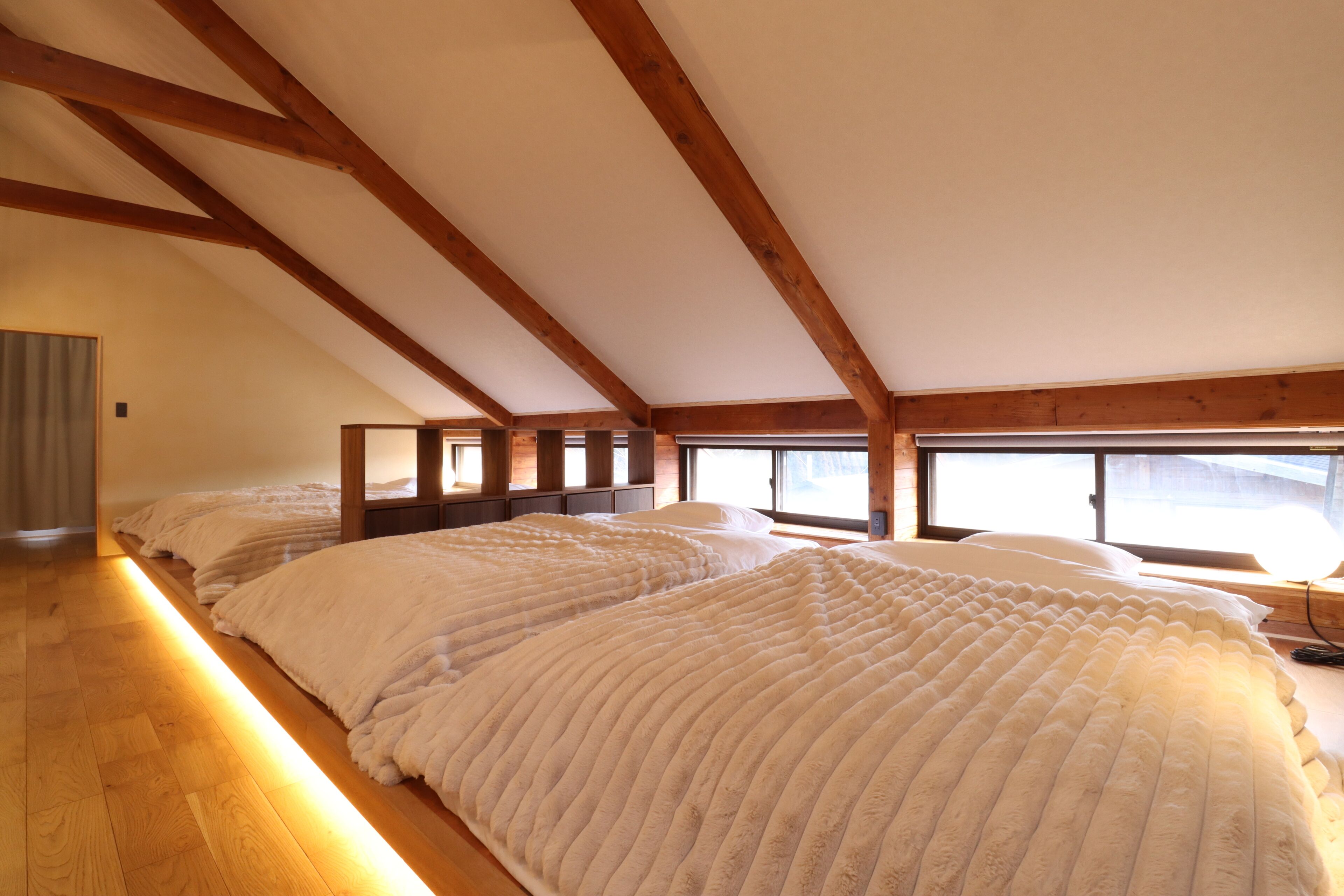 Um quarto espaçoso no sótão, com vigas de madeira, janelas amplas e uma cama aconchegante com manta texturizada.
