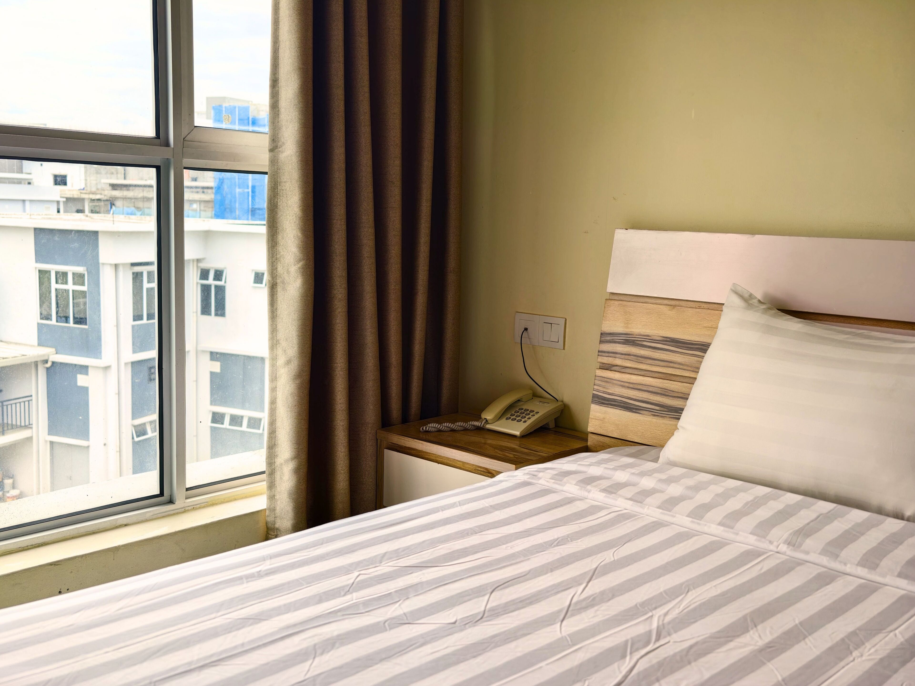 Quarto de hotel com uma cama, mesa de cabeceira com telefone, janela com cortinas e vista para edifícios.