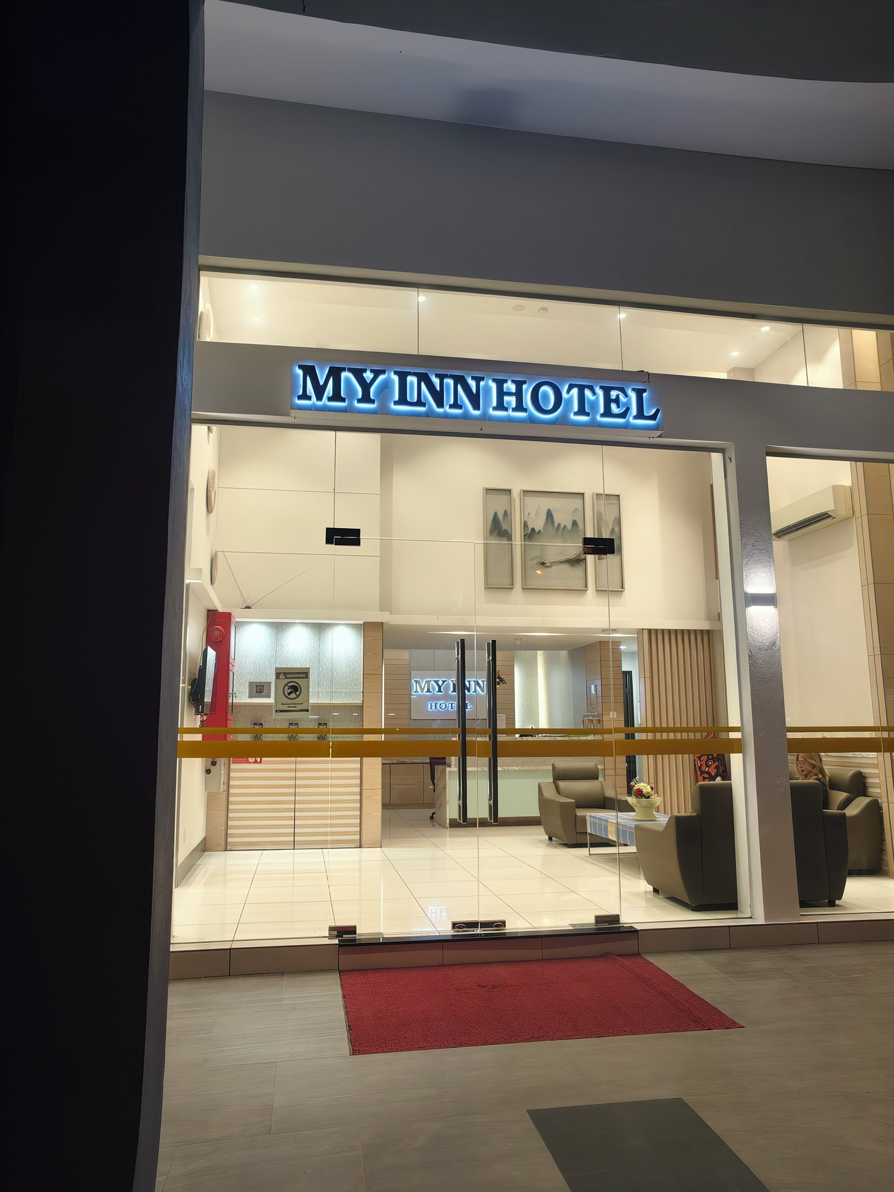 Entrada de hotel com balcão de recepção, área de estar e um letreiro indicando 'MY INN HOTEL'.