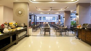 Ampla área de refeições de hotel com várias mesas e cadeiras, um balcão de buffet com alimentos e vista para a paisagem urbana através de grandes janelas.