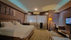 Quarto de hotel com uma cama grande, uma escrivaninha, uma cadeira, uma televisão e uma luminária.