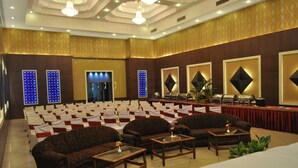 Sala de conferência com fileiras de cadeiras brancas, palco e área de estar com sofás e mesas.