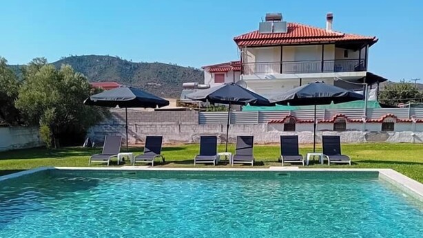 Uma piscina límpida com espreguiçadeiras e guarda-sóis, um edifício com telhado vermelho ao fundo e uma paisagem acidentada.