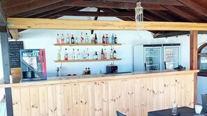 Uma área de bar rústica ao ar livre, com móveis de madeira e uma estante exibindo diversas garrafas.
