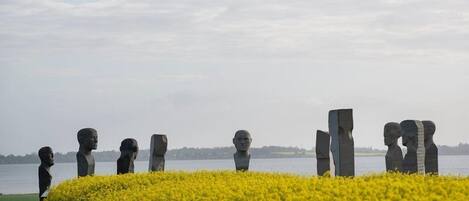 Fileira de estátuas de pedra em um campo de flores amarelas, com um corpo d'água e céu nublado ao fundo.